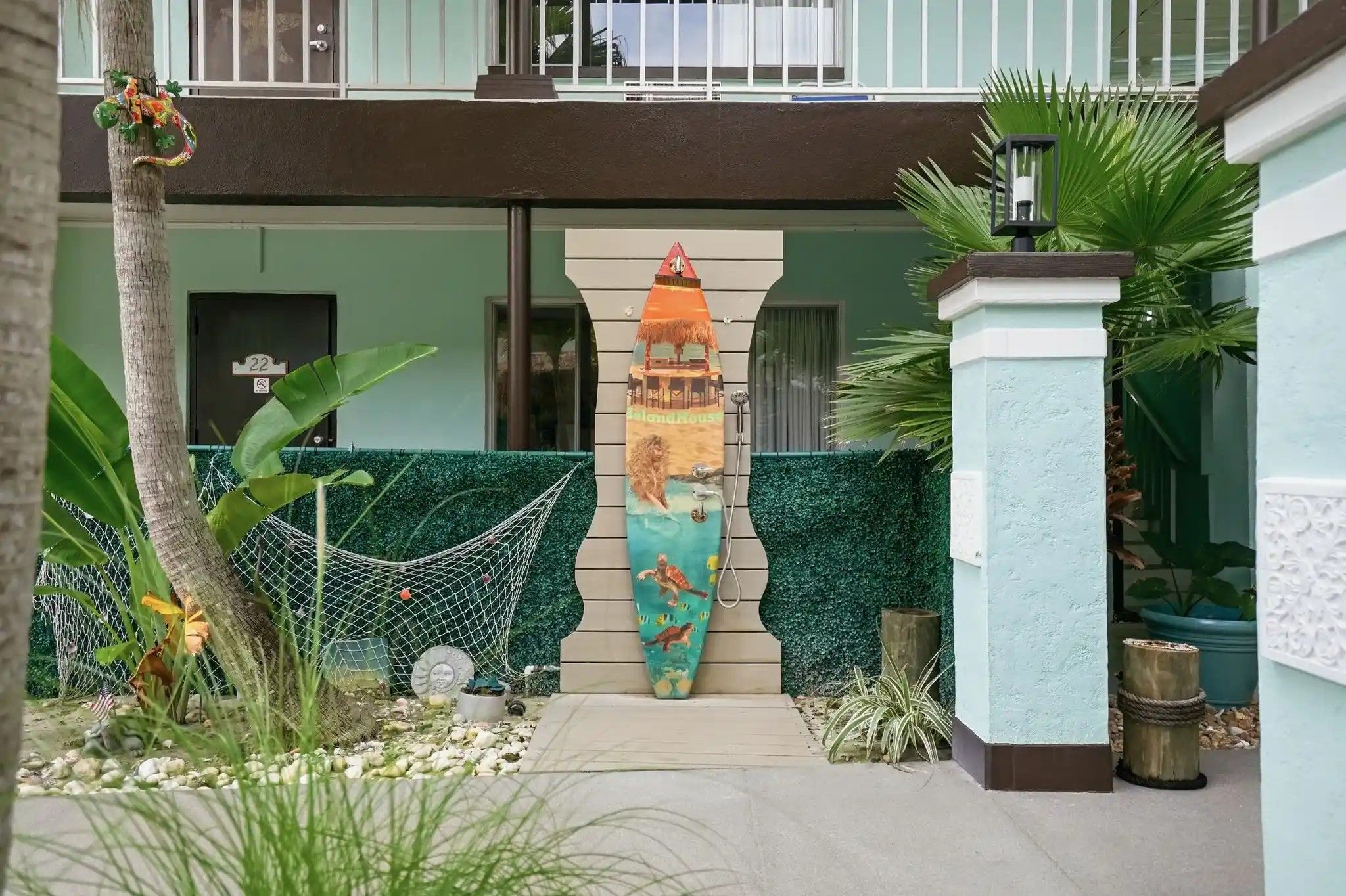 Entrada com tema tropical, apresentando um grande mural, área cercada com plantas e um edifício com varanda.