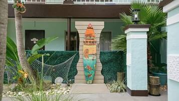 Entrada com tema tropical, apresentando um grande mural, área cercada com plantas e um edifício com varanda.