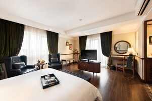 Quarto de hotel com uma cama grande, duas poltronas, uma escrivaninha com televisão e um espelho.