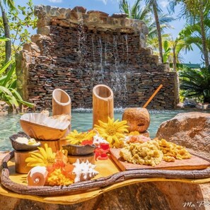 Cenário tropical com uma cascata de pedra, bandeja de madeira com comida e bebidas, e palmeiras ao fundo.