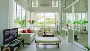 Sala de estar moderna com sofá, mesa de centro e televisão. Grandes janelas oferecem iluminação natural e vista para o verde lá fora.