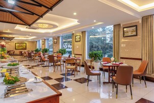Um espaço de refeições de hotel com várias mesas e cadeiras, um buffet com diversos pratos e uma grande janela com vista para árvores.