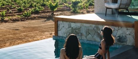 Duas mulheres sentadas à beira de uma piscina, com um vinhedo ao fundo.
