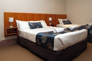 Quarto de hotel com duas camas, cada uma com cabeceira de madeira, roupa de cama branca e colcha estampada escura. Há mesas de cabeceira com abajures e uma tomada na parede.