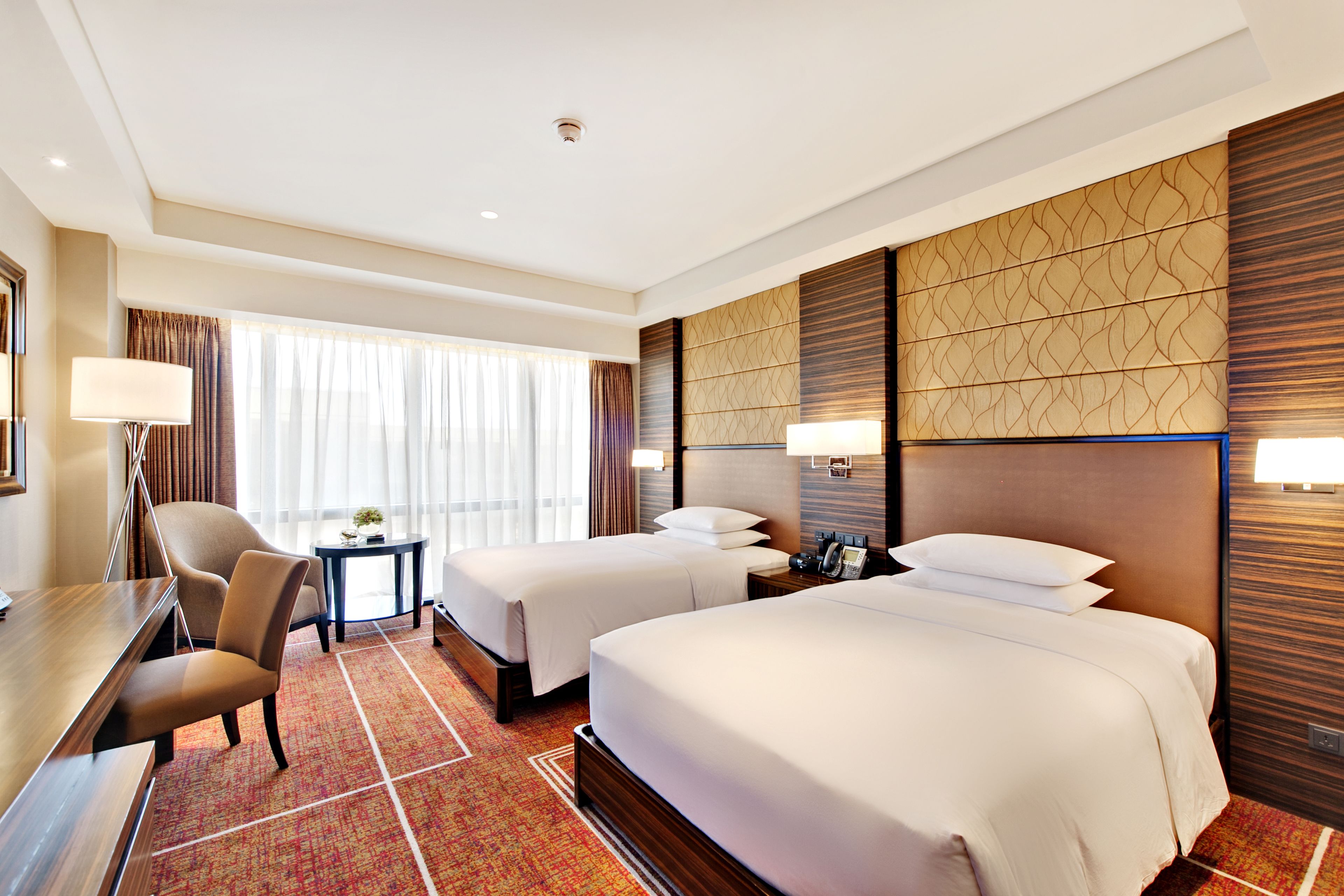 Quarto de hotel com duas camas, uma escrivaninha, uma cadeira e uma mesinha.