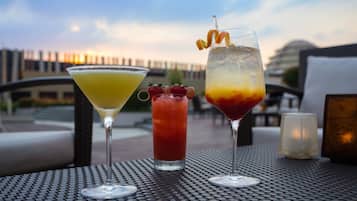 Três cocktails sobre uma mesa, com vista para um pôr do sol e edifícios ao fundo.