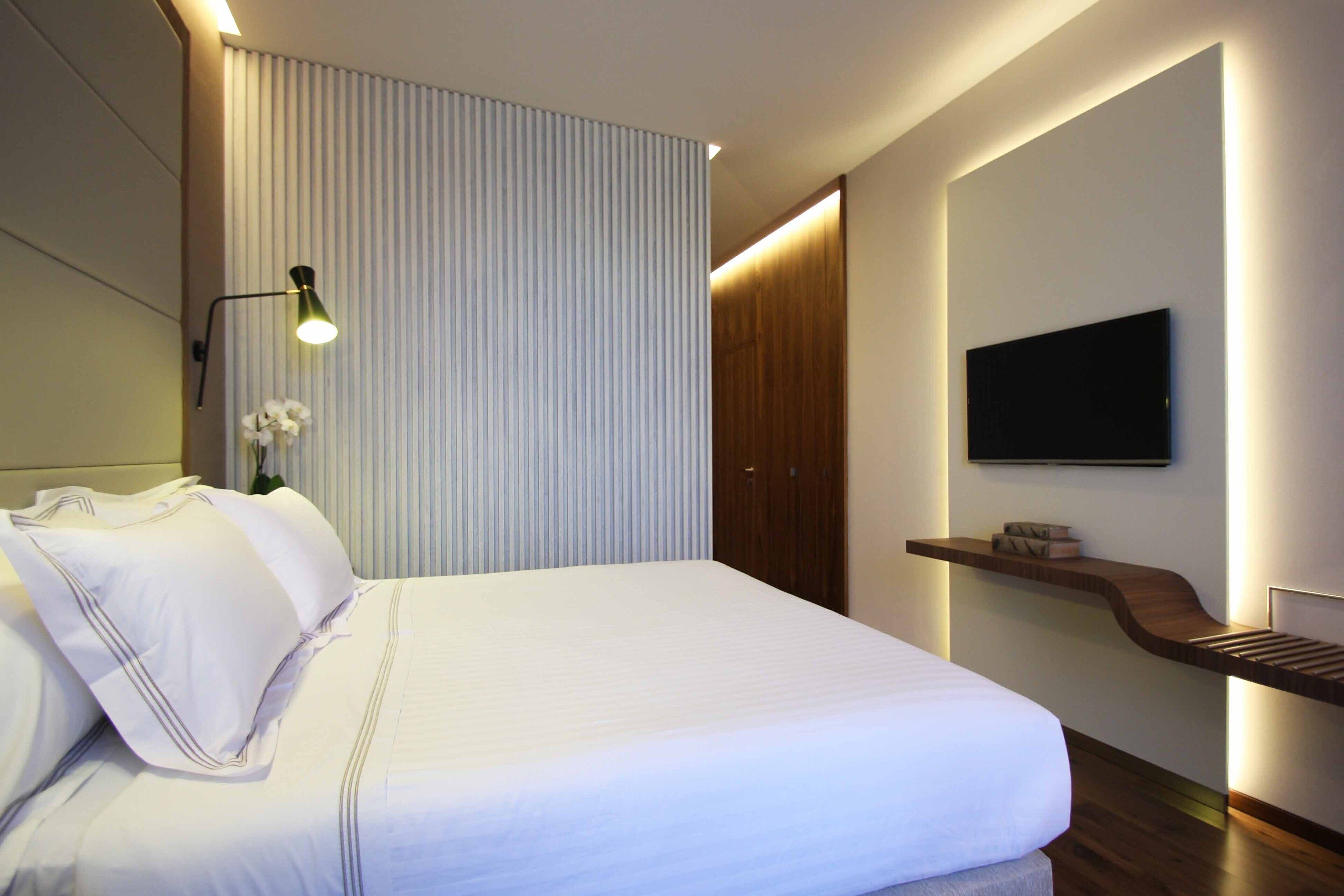 Quarto de hotel moderno com uma cama grande, uma luminária de cabeceira, uma TV montada na parede e uma prateleira de madeira.