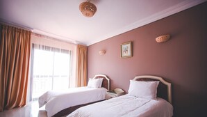 Quarto de hotel com duas camas, uma janela grande com cortinas, um lustre no teto e um quadro na parede.
