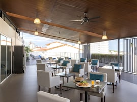Uma área de refeições no terraço com várias mesas e cadeiras, um ventilador de teto e vista para a cidade.