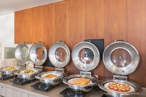 Buffet com diversos alimentos em recipientes de aço inoxidável, em frente a uma parede de madeira.