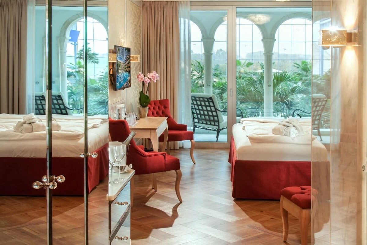Quarto de hotel com duas camas, área de refeições e grandes janelas com vista para o verde.