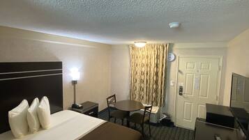 Quarto de hotel com cama, mesa de cabeceira, abajur, escrivaninha, cadeira e porta com relógio.