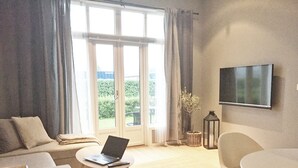 Uma sala de estar moderna com sofá, mesa de centro com um laptop, TV fixada na parede e vista para o exterior através de uma porta de vidro.