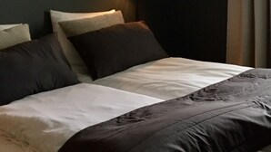 Cama com edredom branco e tons escuros, duas almofadas e luminária fixada na parede.