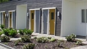 Fileira de cabanas individuais com portas separadas, um caminho pavimentado e pequenas plantas em primeiro plano.