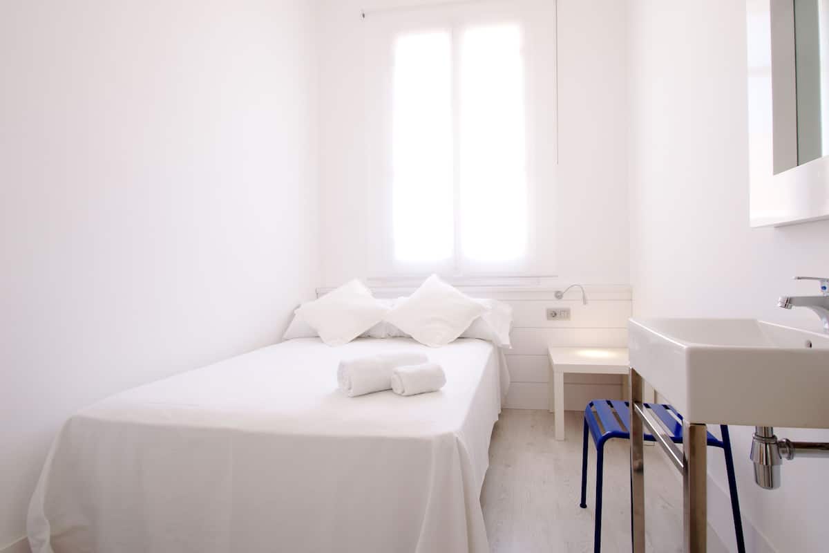 Quarto de hotel claro e branco, com uma cama, uma pequena mesa de cabeceira, um criado-mudo, um banquinho azul e uma pia branca.