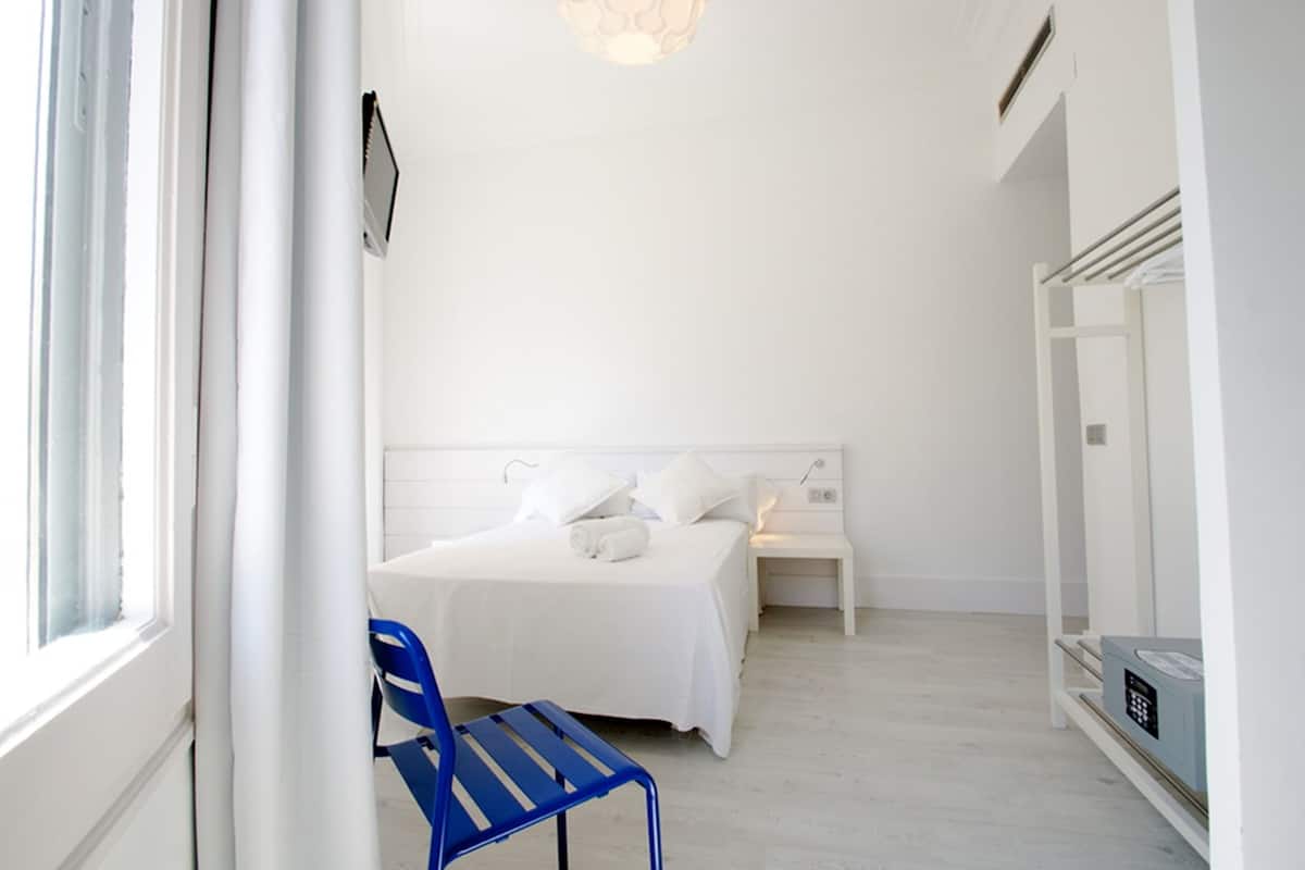 Um quarto branco com uma cama, uma cadeira azul, uma mesa de cabeceira com abajur e um guarda-roupa.