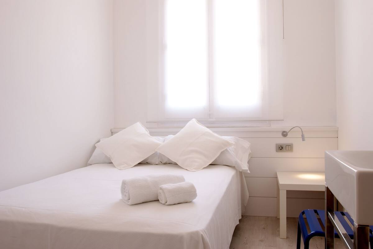 Quarto branco com uma cama, uma mesinha de cabeceira e uma janela com persianas.