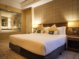 Quarto de hotel com uma cama grande, mesa de cabeceira, espelho e cabeceira com painel de luz.