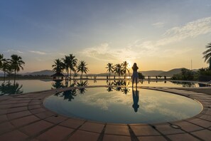 Uma piscina infinita circular com palmeiras e vista para montanhas ao pôr do sol.