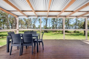 Área externa coberta com deck de madeira, mesa e cadeiras, e vista para árvores e água.