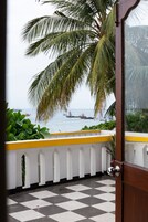 Uma varanda com piso xadrez, vista do mar com barcos e uma palmeira.