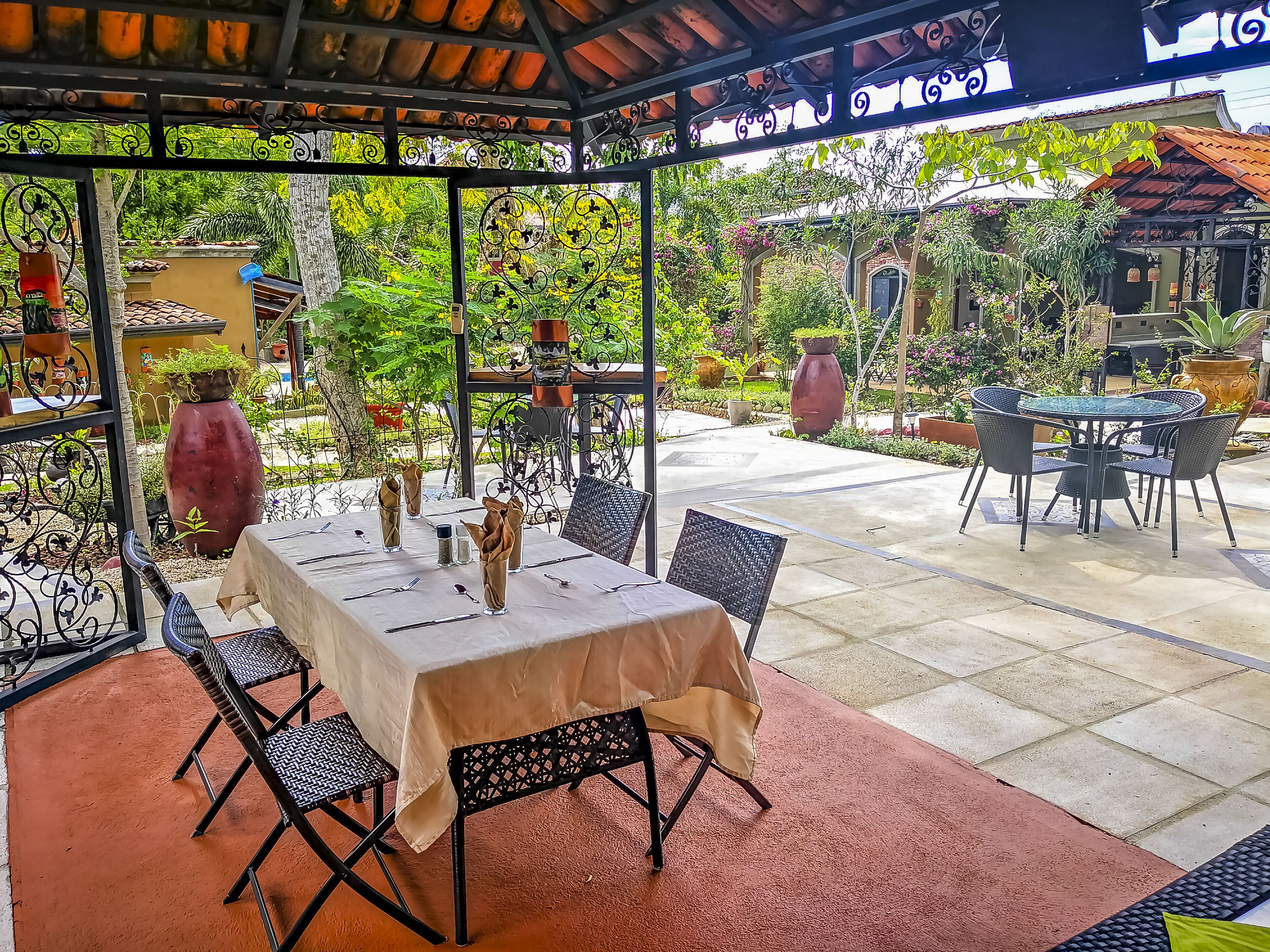 Área de refeições ao ar livre com um terraço coberto, mesas preparadas com talheres e cadeiras.