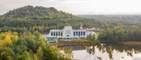 Um edifício moderno com fachada simétrica, cercado por um lago e uma floresta densa.