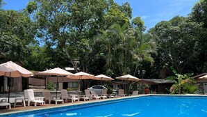 Piscina com espreguiçadeiras e guarda-sóis, cercada por árvores e um edifício.
