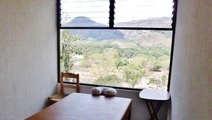 Quarto com mesa de madeira, uma cadeira, um banquinho pequeno e janela com vista para uma paisagem montanhosa.