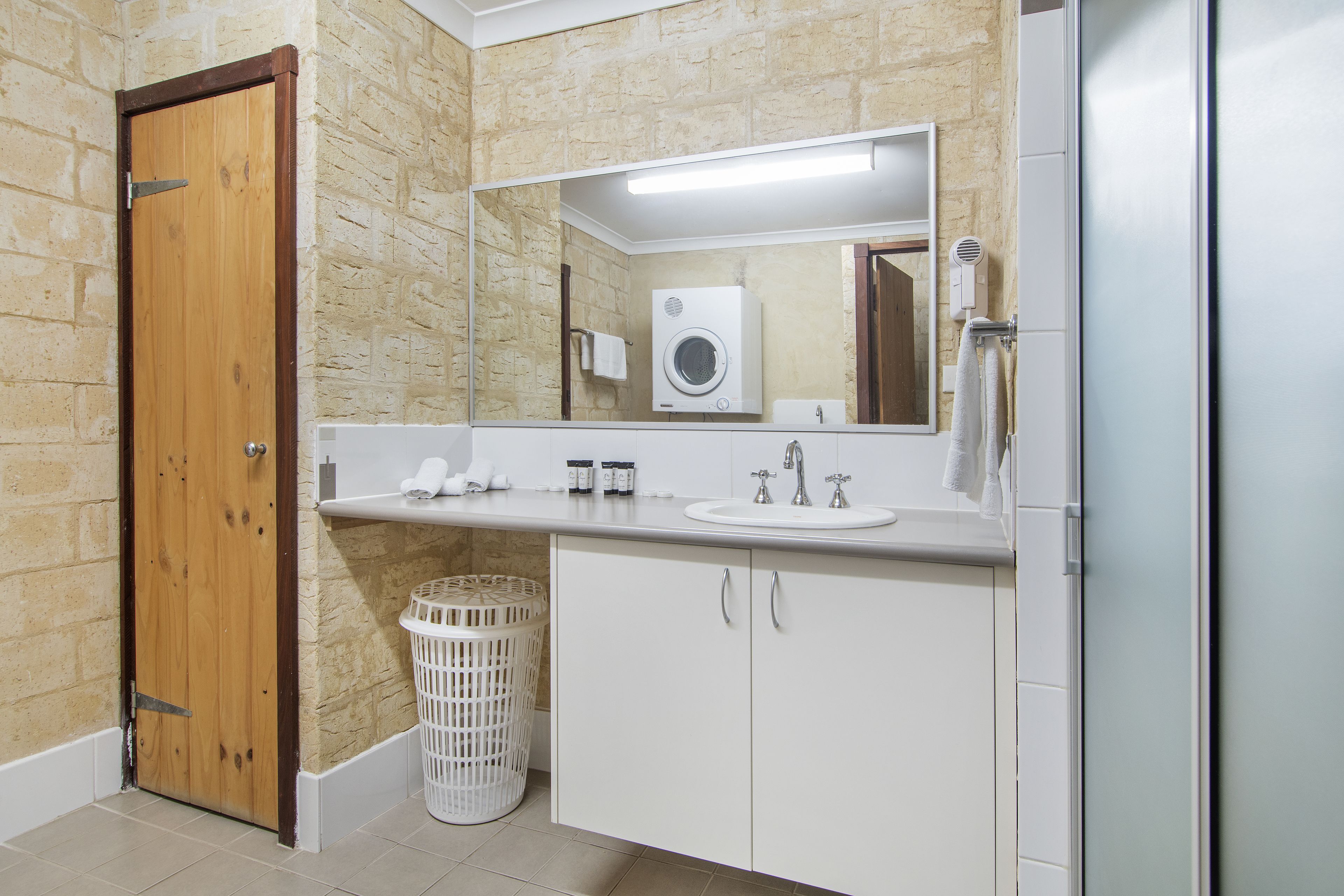 Uma casa de banho com porta de madeira, uma pia branca, um espelho, uma máquina de lavar e uma cesto de roupa.