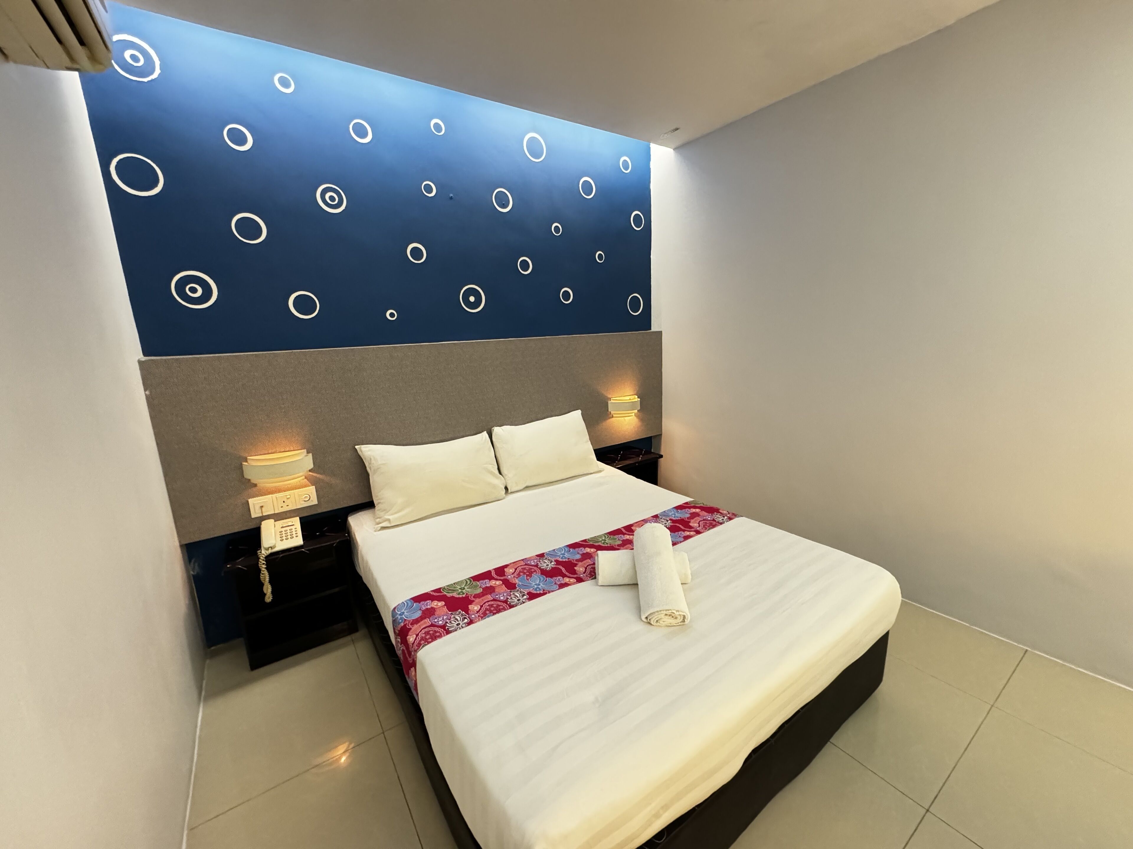 Quarto de hotel moderno com uma cama grande, cabeceira com padrões circulares, luminárias laterais e um telefone na mesa de cabeceira.