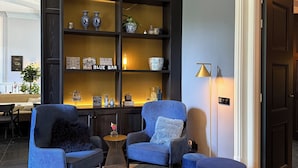 Uma sala de estar com uma estante de madeira escura, dois poltronas azuis, uma mesa de centro redonda e um tapete branco.