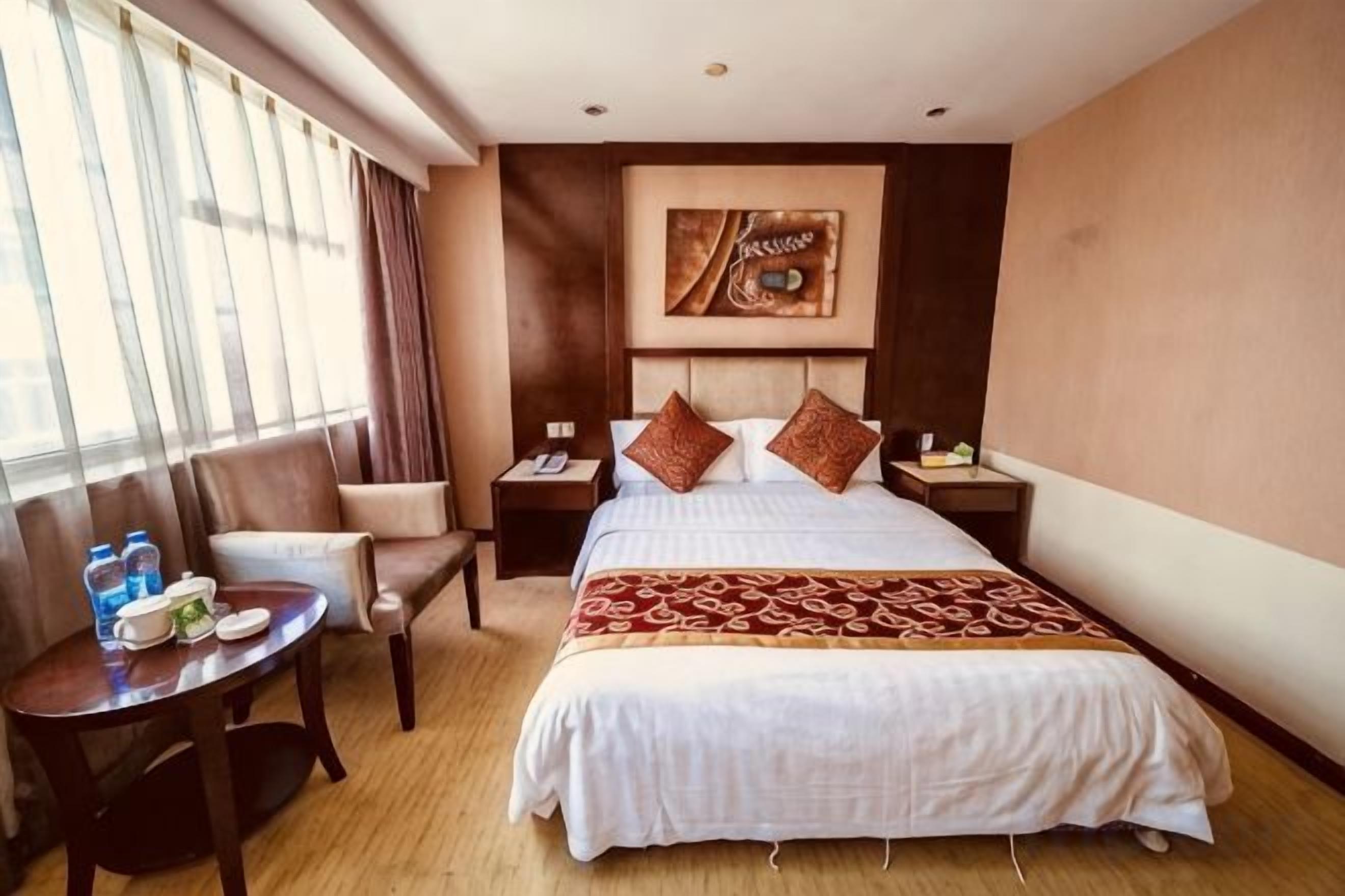 Quarto de hotel com uma cama grande, uma escrivaninha com cadeira, uma mesinha de cabeceira e uma obra de arte na parede.