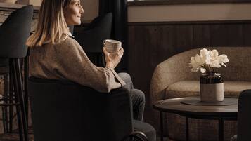 Uma pessoa sentada em uma cadeira, com uma bolsa no chão, uma mesa com um vaso de flores e uma janela com vista para a neve.
