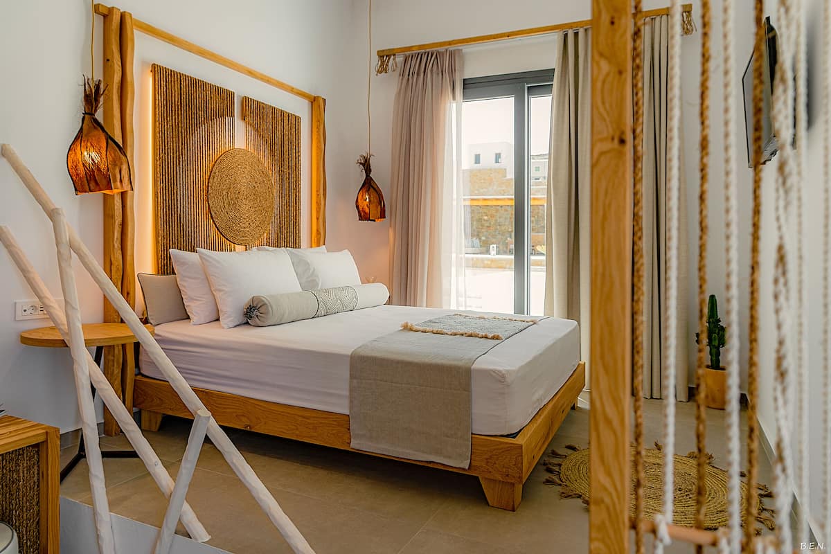 Quarto com cama de madeira, roupa de cama branca, cabeceira trançada e vista da cidade através de uma grande janela.