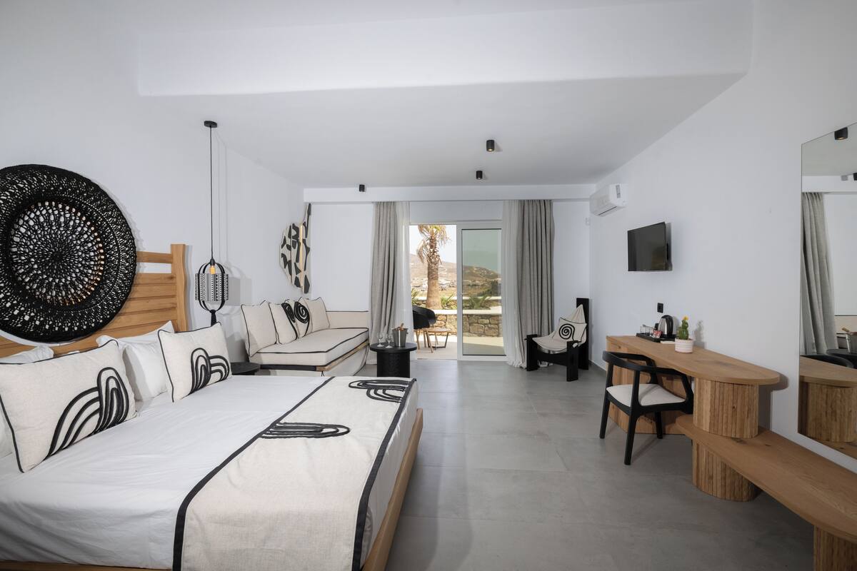 Quarto de hotel moderno com uma cama grande, uma escrivaninha de madeira, um sofá e uma mesa de jantar. Há uma janela ampla com vista para palmeiras e uma varanda.