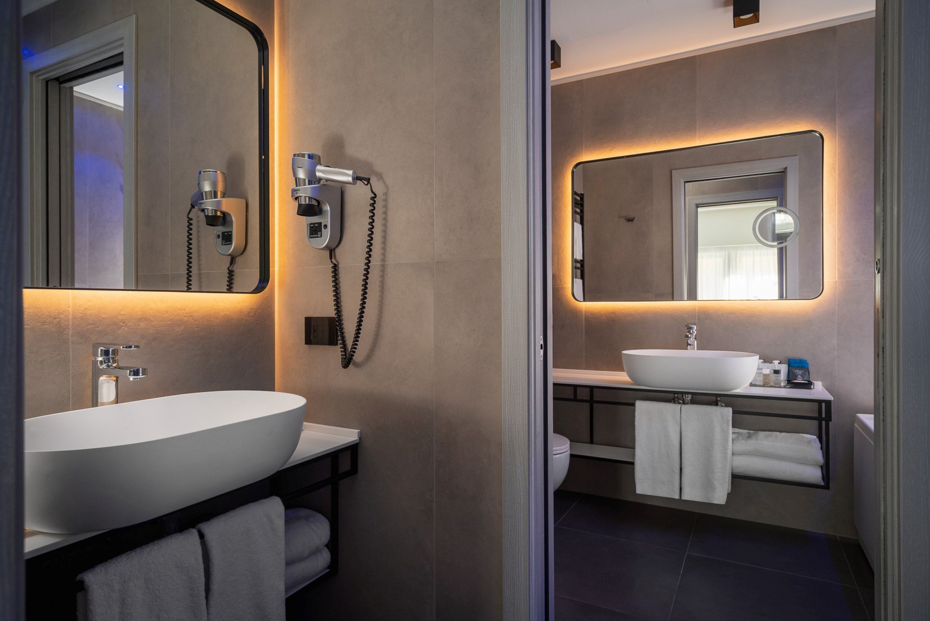 Uma casa de banho moderna com uma grande lavatório oval, um espelho com iluminação integrada, um secador de cabelo e um porta-toalhas.