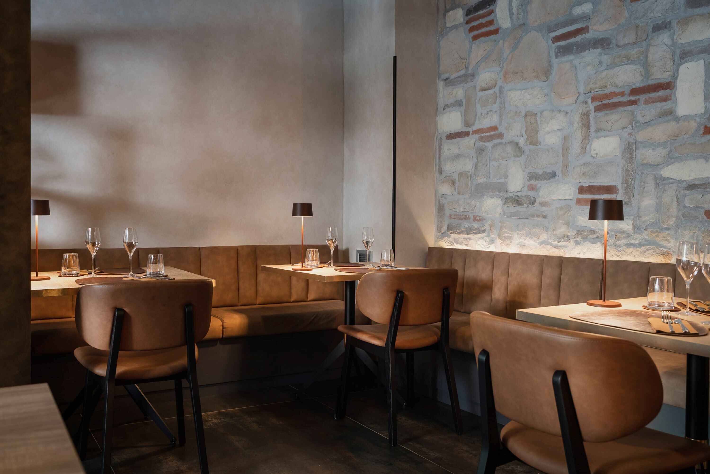 Interior de restaurante com assentos em camarotes, mesas e parede de pedra.