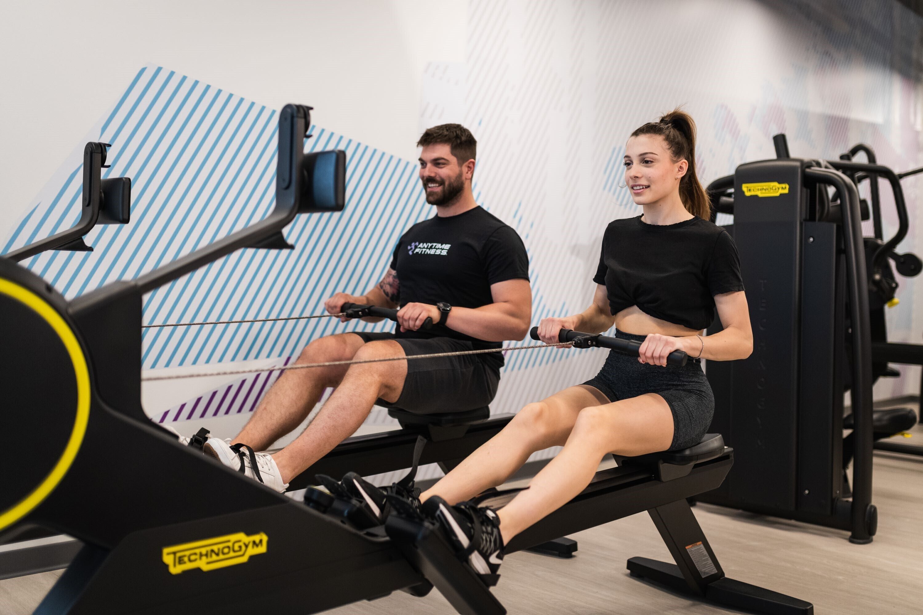 Duas pessoas usando uma máquina de remo em uma academia com o logotipo Technogym.