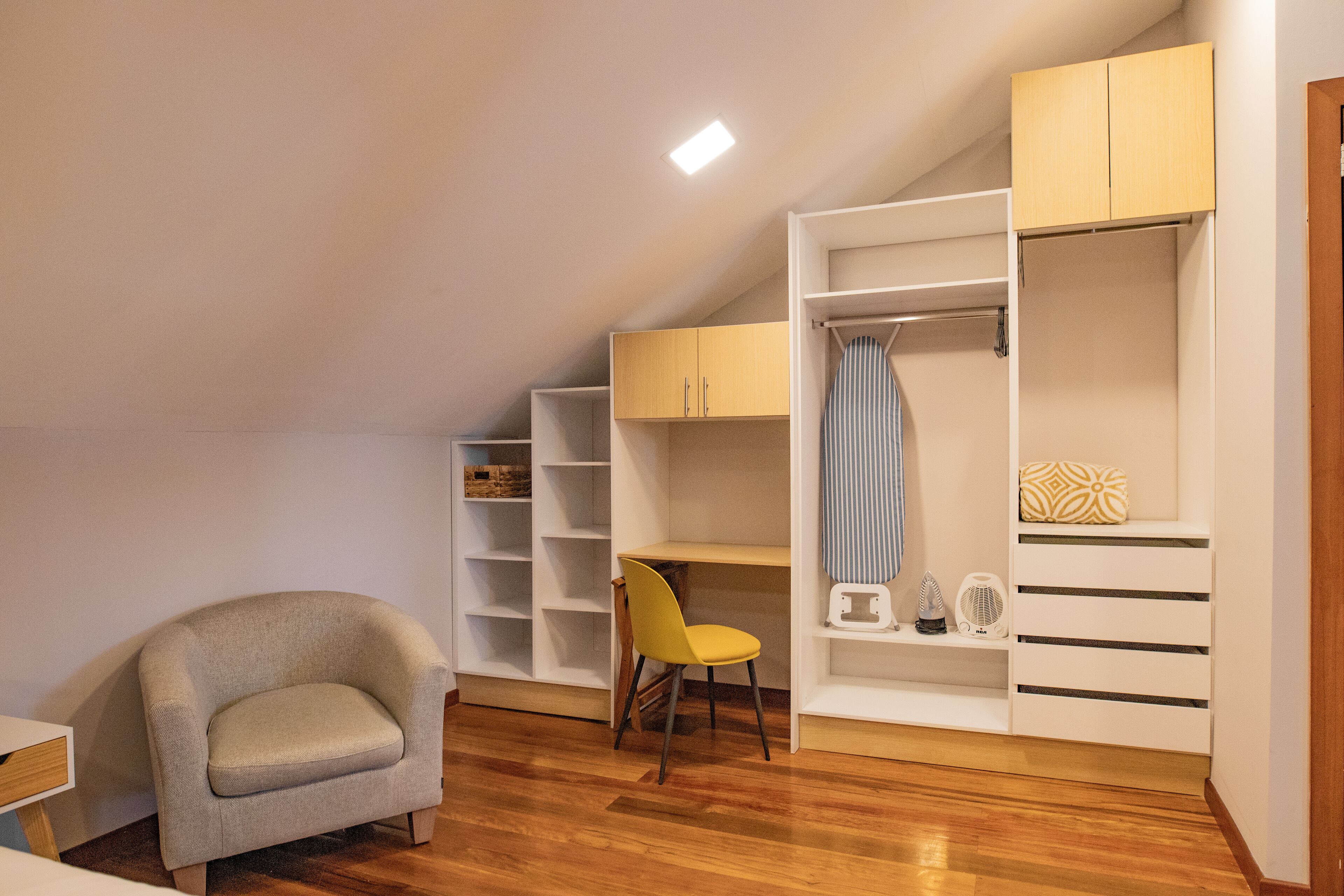 Quarto com piso de madeira, poltrona cinza, cadeira amarela, escrivaninha e guarda-roupa embutido com prateleiras e espaço para pendurar.