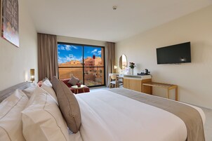 Quarto de hotel com uma cama grande, uma escrivaninha, uma cadeira, uma televisão e vista para edifícios através de uma janela ampla.