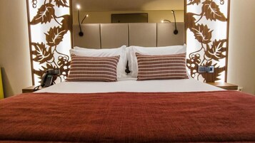 Quarto de hotel com uma cama grande, duas almofadas decorativas, telefone ao lado da cama e luminária fixada na parede.