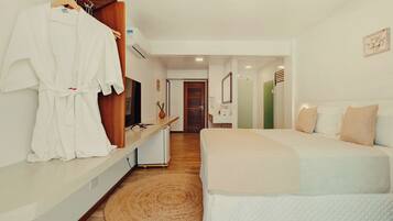 Quarto de hotel com cama, um banco de madeira, roupão pendurado em um cabide, televisão e espelho.