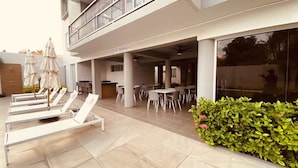 Uma área externa moderna com espreguiçadeiras, um terraço com mesas e cadeiras, e uma placa com a inscrição
