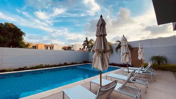 Piscina com espreguiçadeiras e guarda-sóis, cercada por um muro baixo e palmeiras.