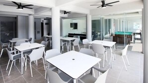 Uma área de refeições interna moderna, com mesas e cadeiras brancas, ventiladores de teto e uma mesa de bilhar ao fundo.