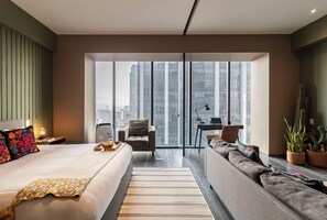 Quarto de hotel moderno com uma cama grande, um sofá, uma mesa com um laptop e vista para a cidade através de grandes janelas.