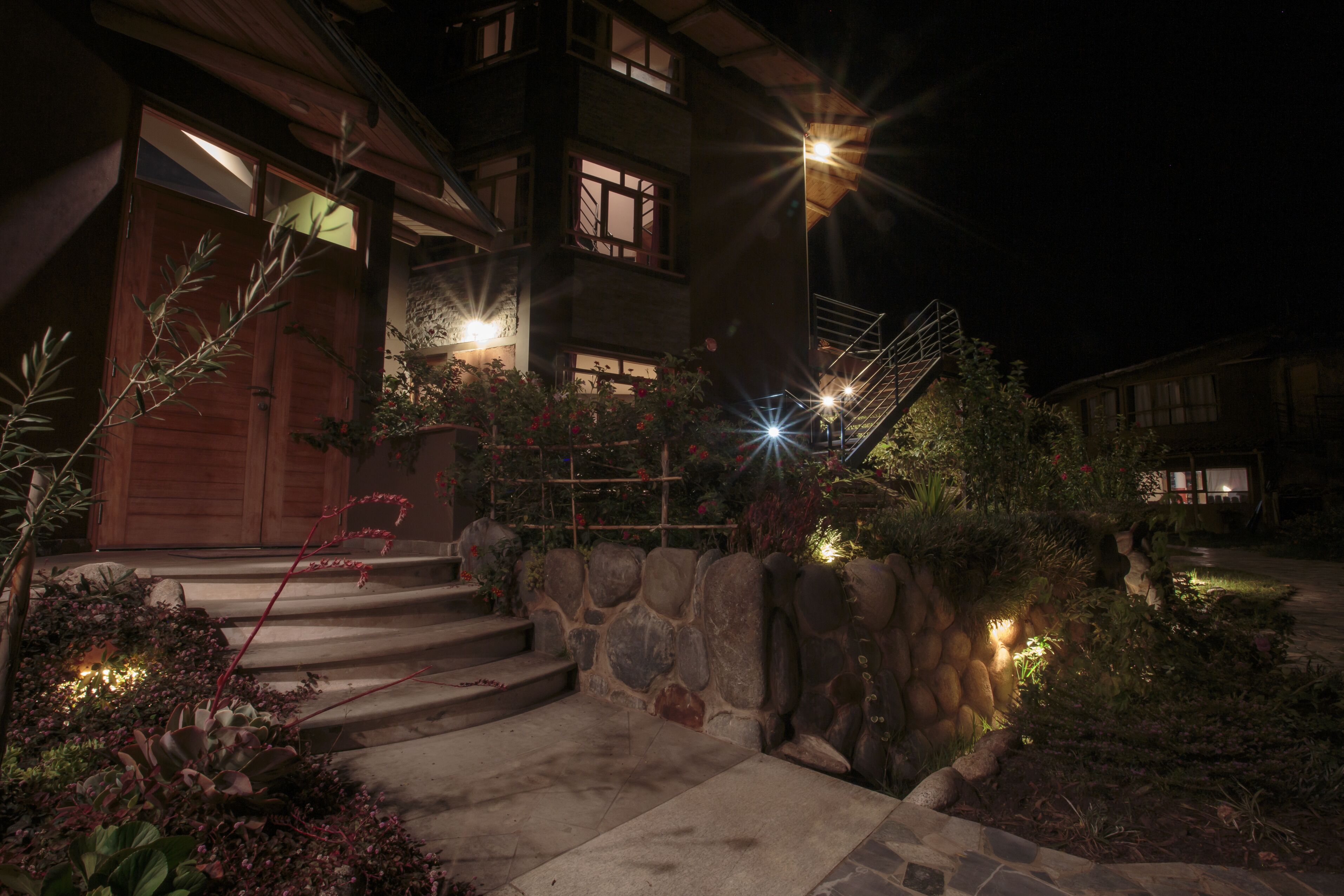 Um edifício de vários andares com uma escadaria de pedra que leva à entrada, cercado por vegetação exuberante e iluminado por luzes externas.
