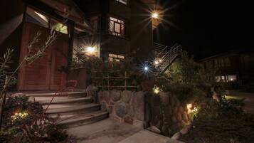 Um edifício de vários andares com uma escadaria de pedra que leva à entrada, cercado por vegetação exuberante e iluminado por luzes externas.